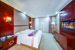 Quarto de hotel com uma cama grande, uma escrivaninha com telefone, uma TV fixada na parede e uma janela com cortinas.