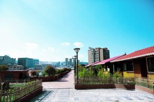 Terraço no telhado com um caminho que leva a um edifício de telhado vermelho, cercado por vegetação e um céu limpo.