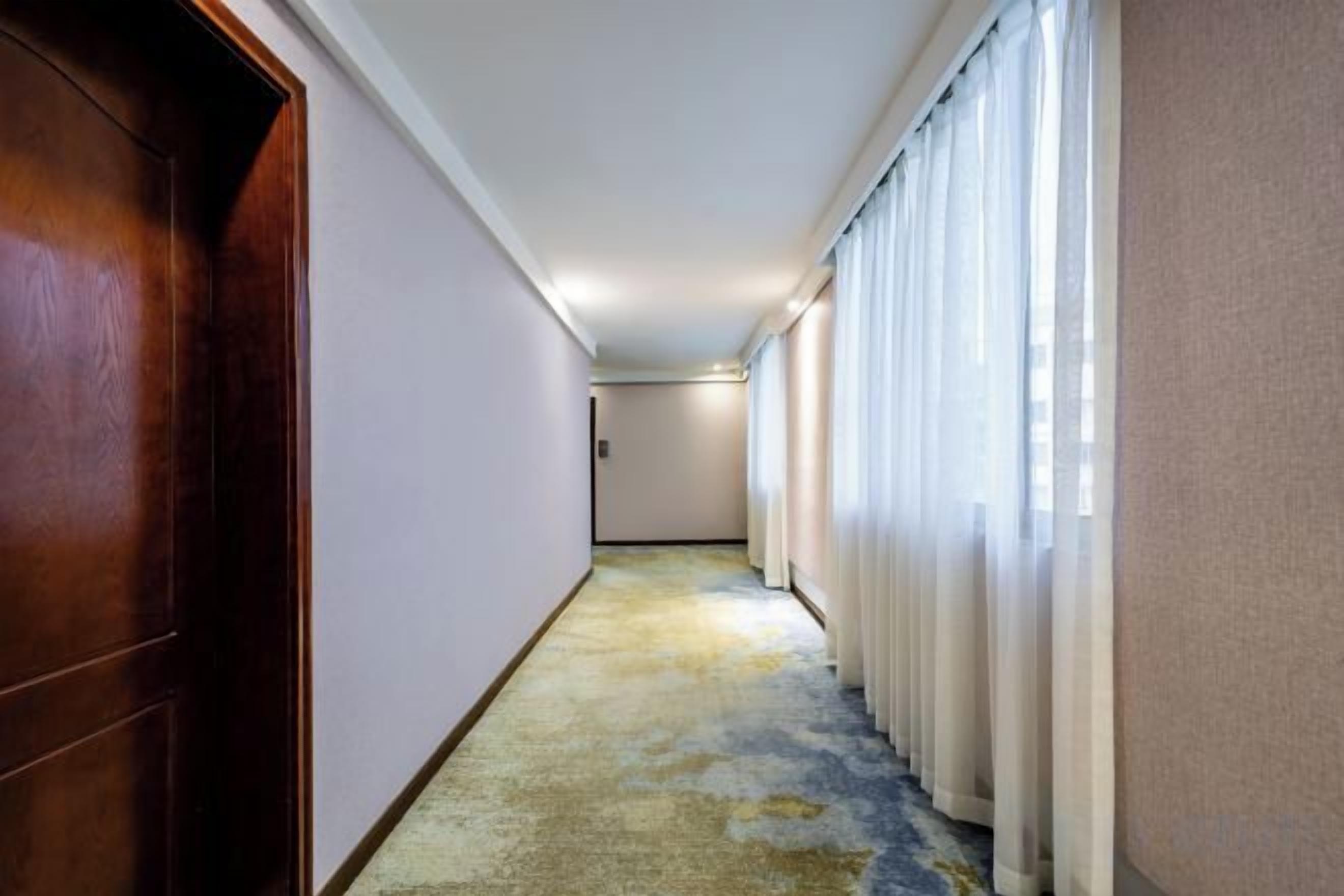 Corredor de hotel com porta de madeira, piso coberto por carpete e janela com cortinas.