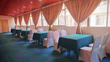 Sala de conferências com mesas, cadeiras, janelas amplas com cortinas e piso coberto por carpete.