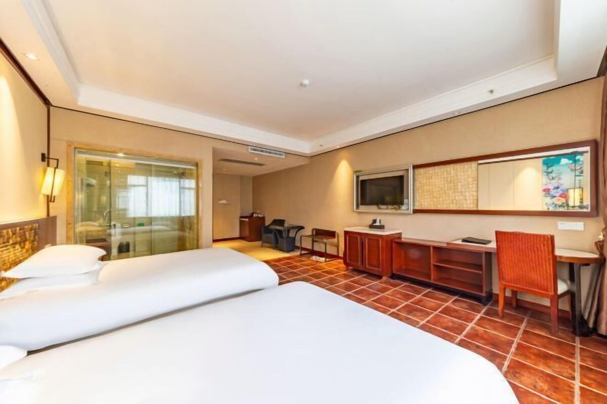 Quarto de hotel com cama, escrivaninha, cadeira, televisão e banheiro com porta de vidro.