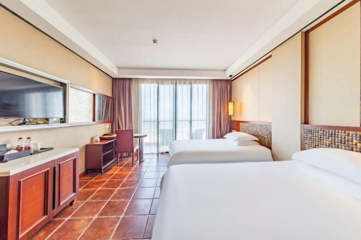 Quarto de hotel com duas camas, uma escrivaninha com cadeira, televisão e janela com cortinas.