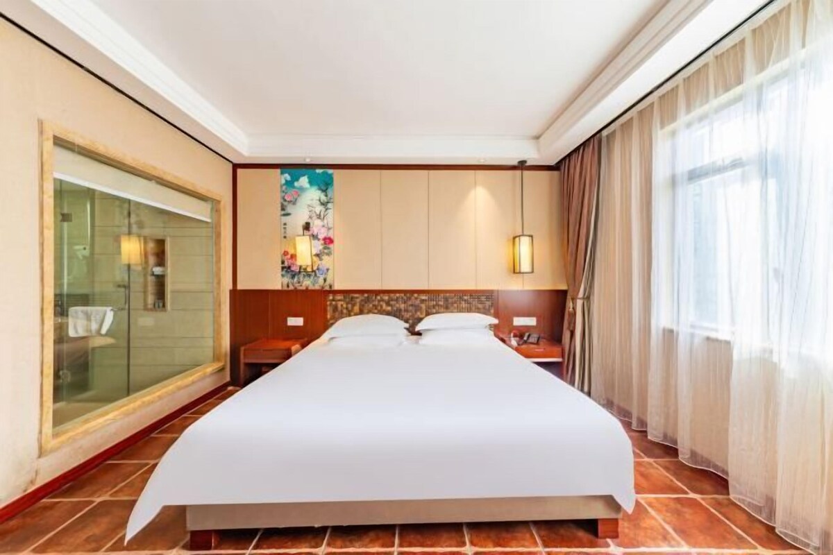 Quarto de hotel com uma cama grande, box de vidro para o chuveiro, um detalhe decorativo na parede e vista para o exterior através de cortinas translúcidas.