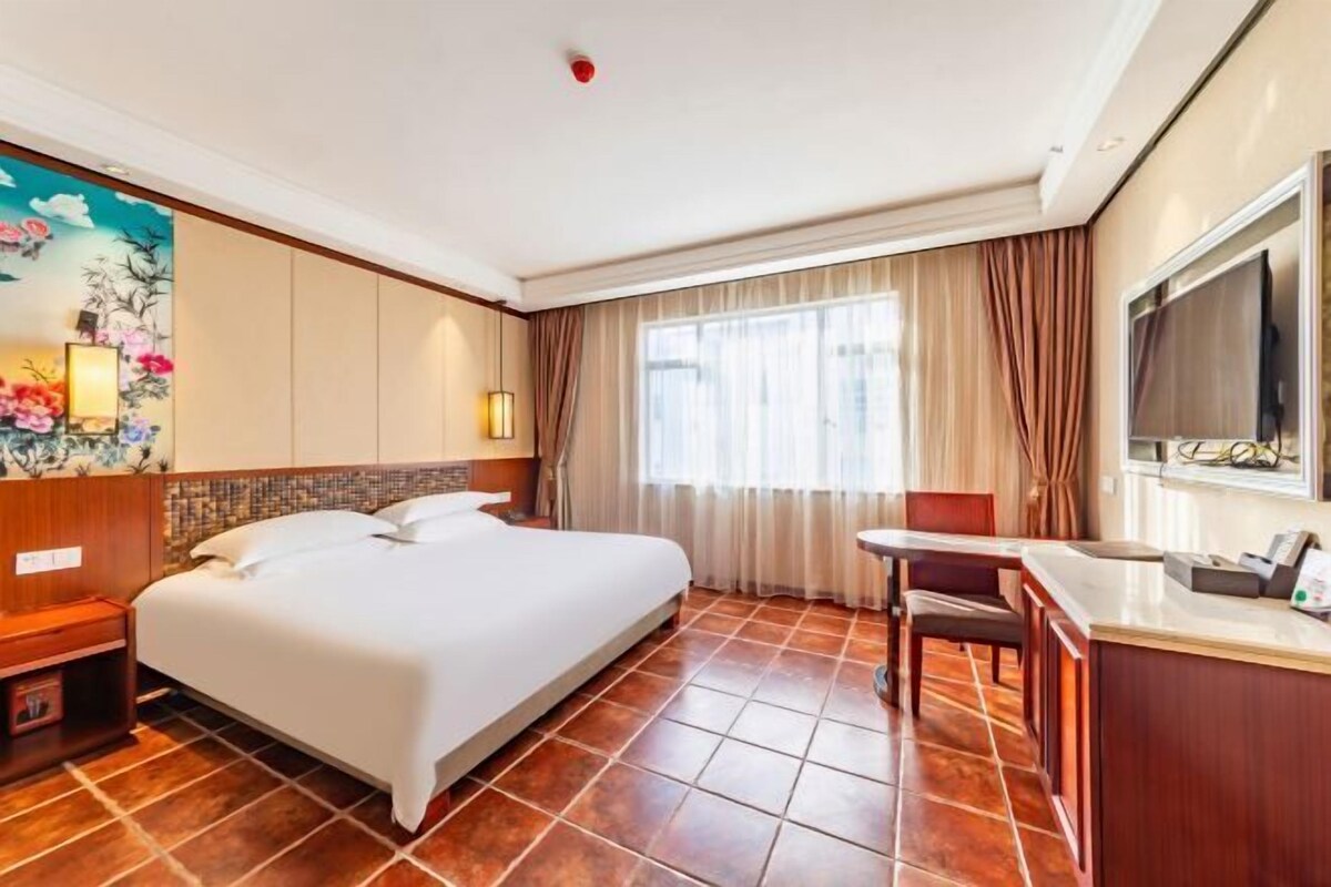 Quarto de hotel com uma cama grande, uma escrivaninha, uma TV e um quadro na parede.
