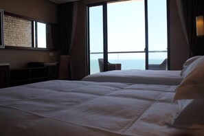 Quarto de hotel com duas camas, uma janela grande com vista para o mar e uma mesinha.