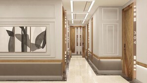 Corredor moderno de hotel com elevador central, uma grande obra de arte abstrata à esquerda e uma porta de madeira com número à direita.