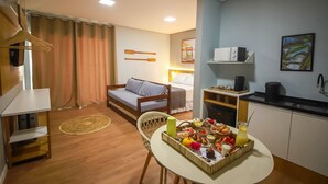 Quarto de hotel compacto com cama, sofá, mesa de jantar com café da manhã, área de cozinha e vista para o exterior.
