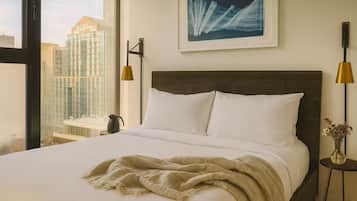 Quarto de hotel com uma cama grande, vista para a cidade, uma pintura abstrata emoldurada e duas luminárias de cabeceira.