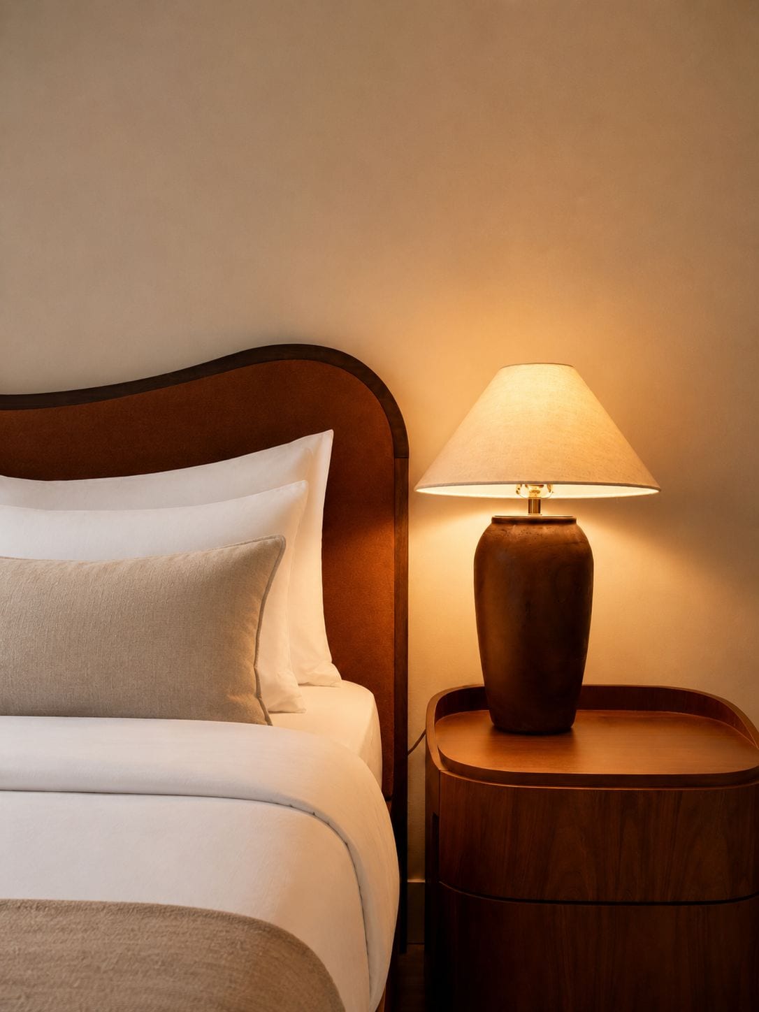Cama bem arrumada com roupa de cama branca e cabeceira de madeira, acompanhada de uma mesinha de cabeceira com abajur.
