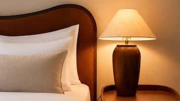 Cama bem arrumada com roupa de cama branca e cabeceira de madeira, acompanhada de uma mesinha de cabeceira com abajur.