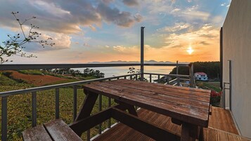 Um deck de madeira com vista para o pôr do sol sobre a água, rodeado por vegetação e montanhas ao longe.