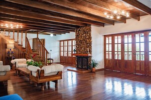 Uma sala de estar ampla, com piso de madeira, lareira de pedra, escada de madeira e grandes janelas.