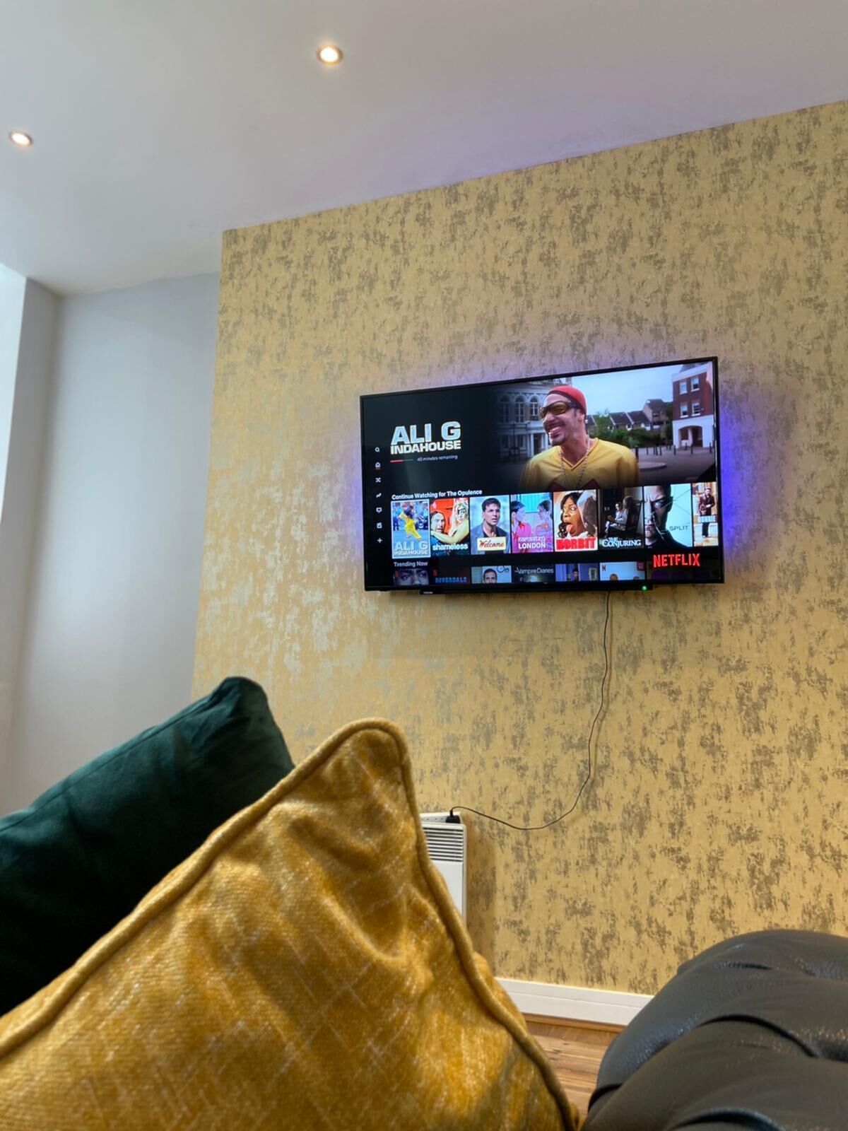 Uma TV de tela plana fixada em uma parede texturizada, exibindo a interface de um serviço de streaming com diversas opções de programas.