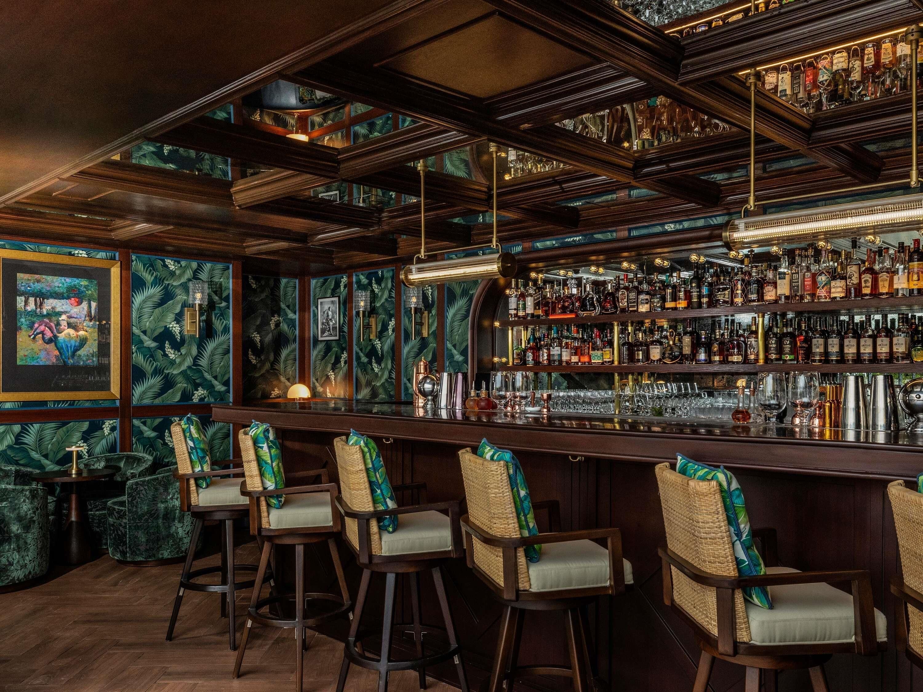 Uma área de bar com banquetas de madeira, uma estante de bebidas bem abastecida e um design de parede com temática tropical.