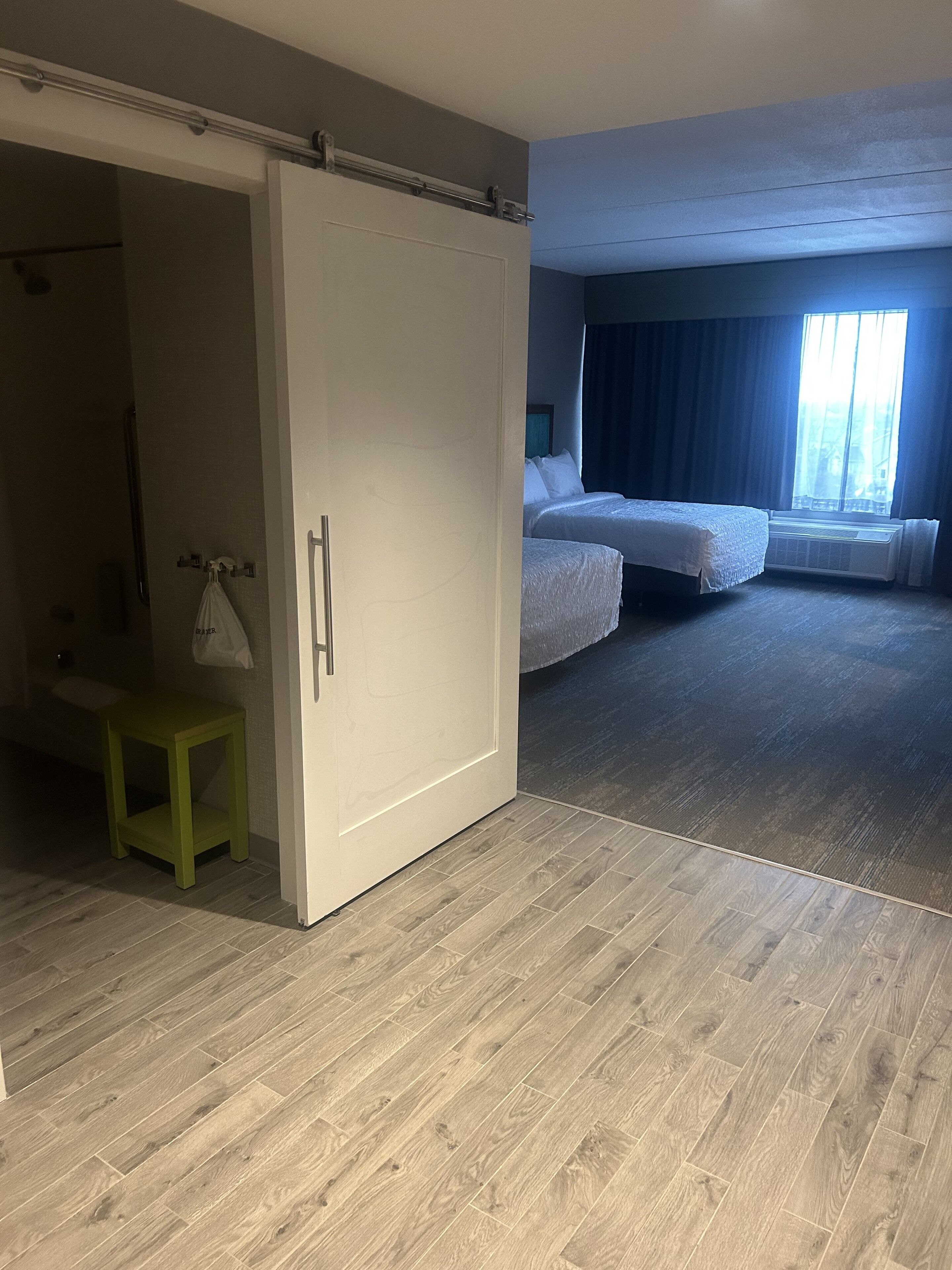 Quarto de hotel com piso de madeira, porta deslizante, cama com roupa branca, um banco e janela com cortinas.