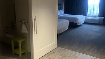 Quarto de hotel com piso de madeira, porta deslizante, cama com roupa branca, um banco e janela com cortinas.