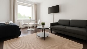 Quarto de hotel moderno com uma cama, um sofá, uma mesa redonda pequena com uma planta, uma televisão na parede e uma janela com cortinas.