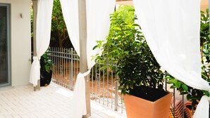 Um espaço ao ar livre coberto, com planta em vaso, cortinas brancas e cerca de metal.