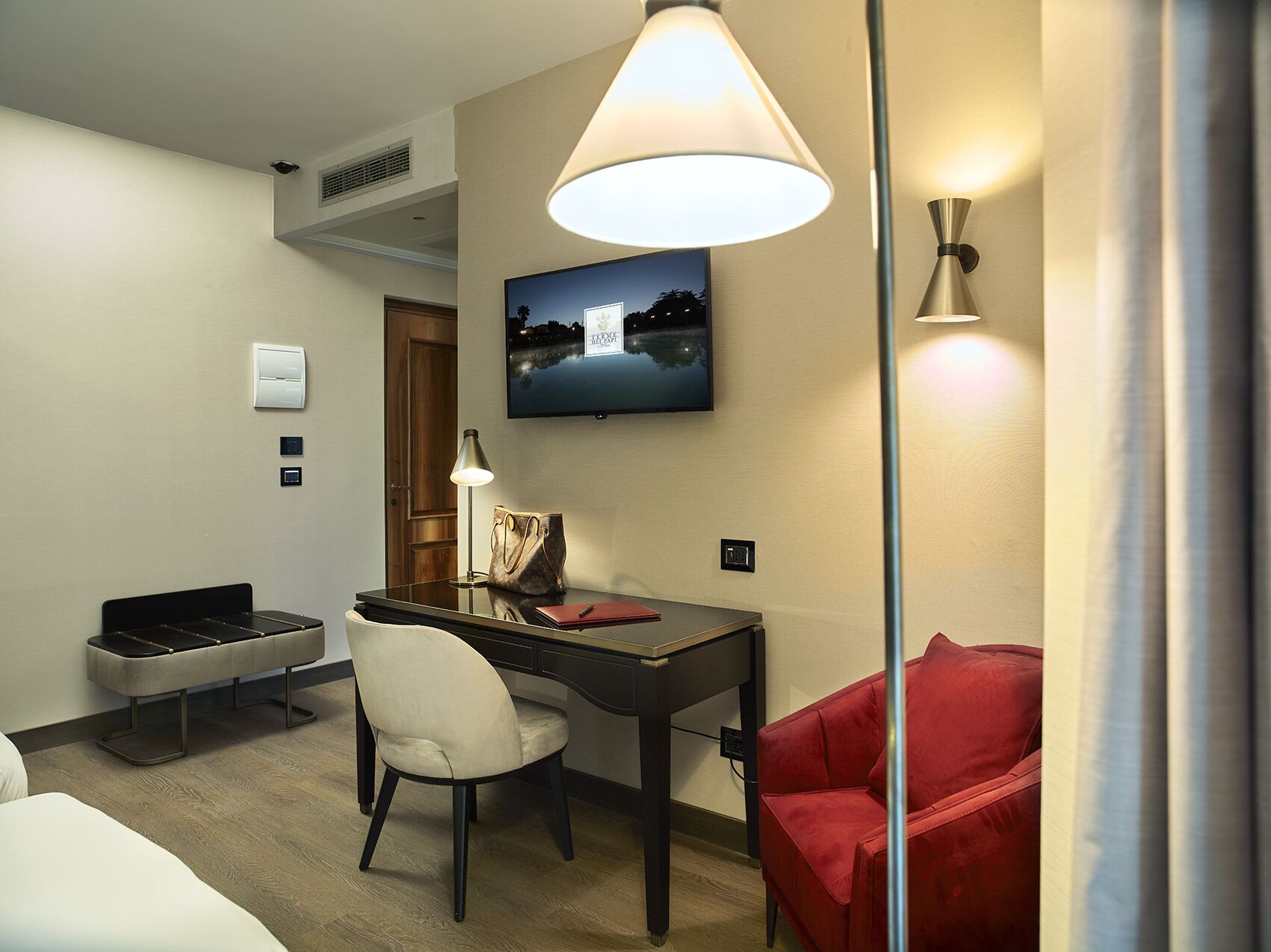 Quarto de hotel com cama, escrivaninha com cadeira, poltrona vermelha, televisão na parede e luminária.