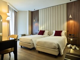 Quarto de hotel com duas camas, uma escrivaninha, uma cadeira e uma mesinha de cabeceira com abajur.