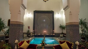 Uma área de piscina interior luxuosa com decoração requintada, incluindo um amplo painel decorativo, sofás vermelhos e plantas em vasos.