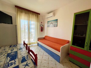 Quarto pequeno com uma cama, uma mesinha, uma cadeira, janela com cortinas, um extintor de incêndio e um armário verde.