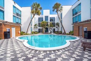 Piscina com deck de azulejos, cercada por palmeiras e edifícios modernos.
