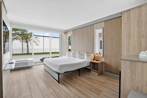 Quarto moderno com uma cama grande, mesa de cabeceira com abajur, espelho e vista para o exterior através de grandes janelas.