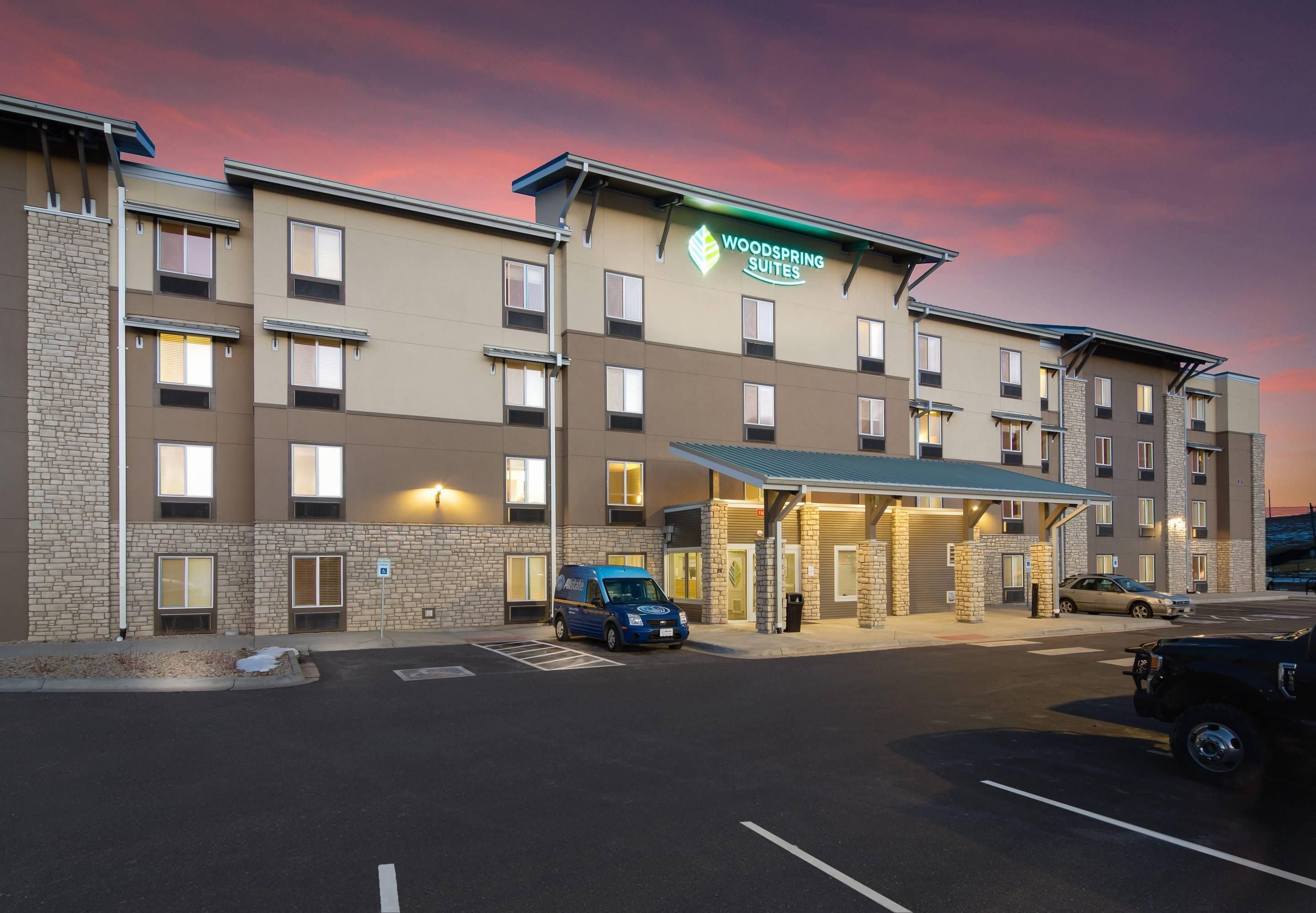 Um hotel de vários andares com telhado plano e uma placa indicando 'Woodspring Suites'. Há várias janelas em cada andar, e a entrada possui uma galeria coberta.
