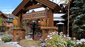 Exterior do Grand Lodge no Peak 7, com entrada de madeira, uma placa, pilares de pedra e plantas cobertas de neve.