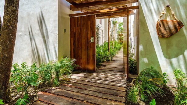 Uma trilha de madeira leva a uma porta de madeira, cercada por vegetação e uma sacola pendurada.