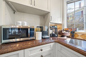 Cozinha com armários brancos, bancada de granito, micro-ondas, cafeteira e janela com vista para árvores.