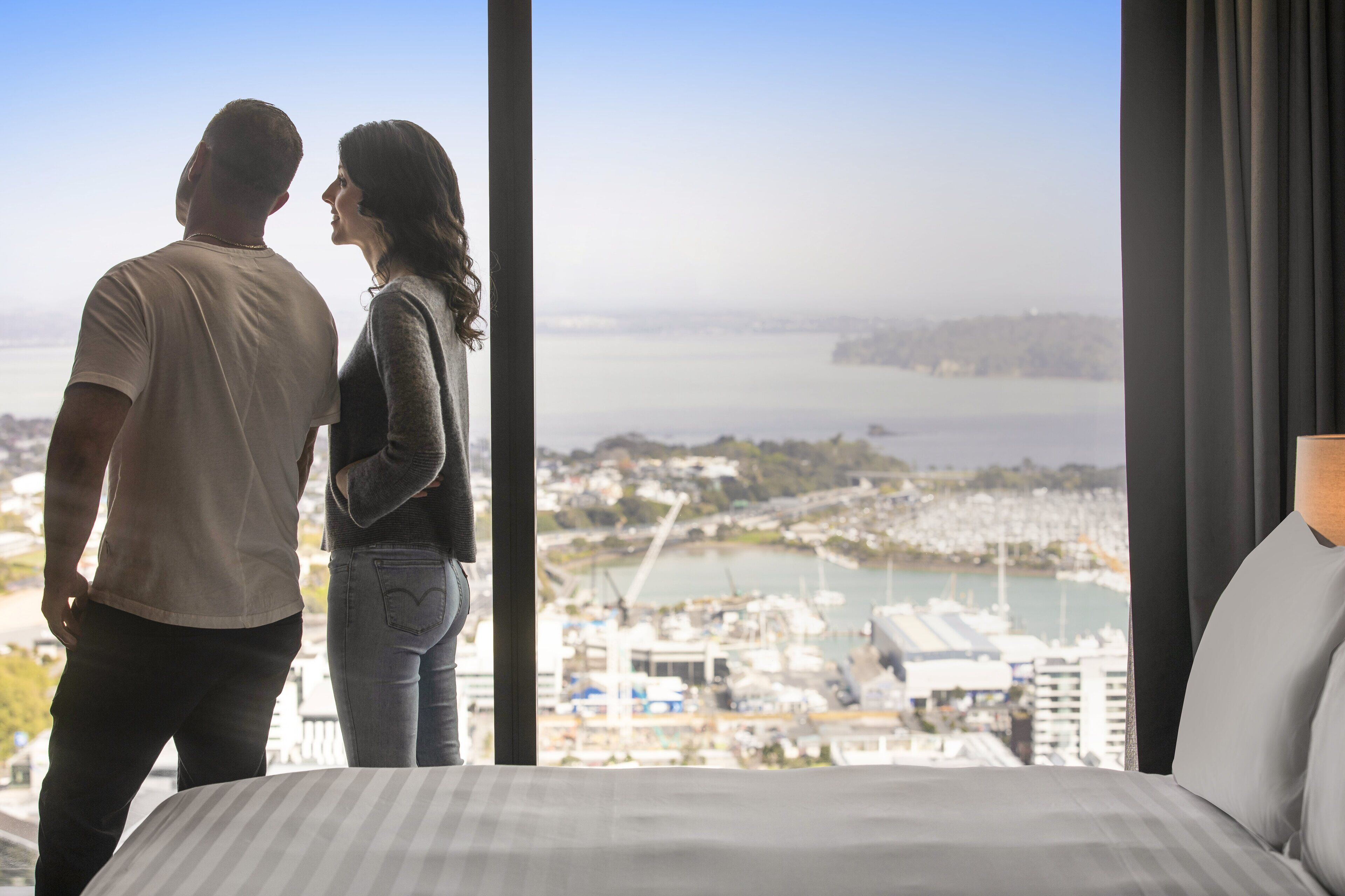 Um casal em pé perto de uma grande janela com vista para um calçadão e a paisagem urbana.
