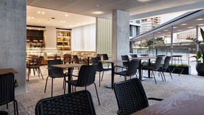 Um café moderno em ambiente interno, com diferentes áreas de estar, incluindo mesas e cadeiras, e uma cozinha à vista com prateleiras repletas de diversos produtos.