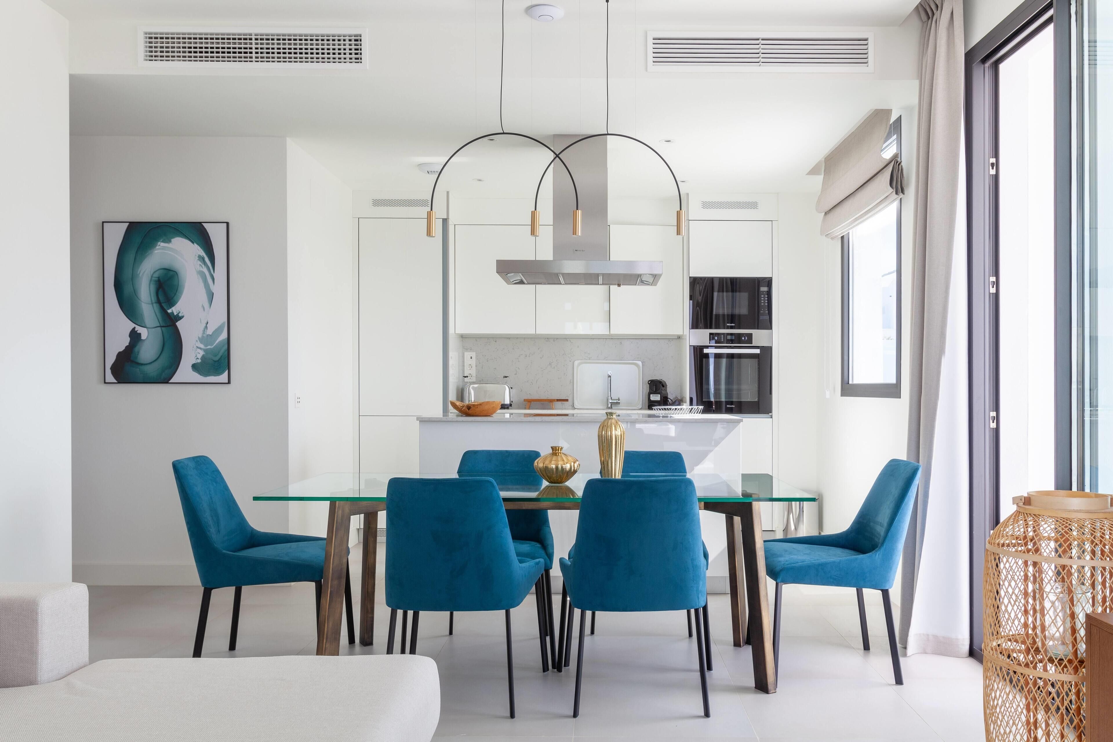 Uma área de refeições moderna com uma mesa de vidro, cadeiras azuis e uma cozinha ao fundo.