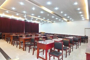 Uma sala de conferências com fileiras de mesas e cadeiras, um palco com cortina vermelha e um quadro branco.
