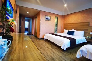 Quarto de hotel com piso de madeira, uma cama grande com roupa de cama branca, uma mesa de cabeceira com telefone e uma televisão fixada na parede.