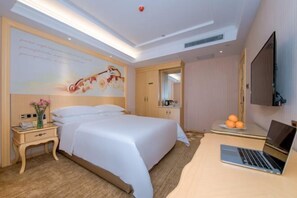 Quarto de hotel com cama, mesa de cabeceira, luminária e um laptop sobre a escrivaninha.