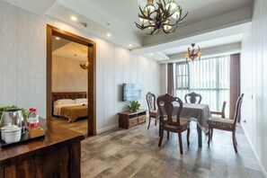 Quarto de hotel com piso de madeira, área de refeições com mesa e cadeiras, TV na parede e um candelabro.