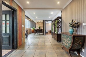 Um corredor de hotel com piso ladrilhado, uma área de refeições com cadeiras e uma mesa, e um aparador decorativo com um vaso de flores.