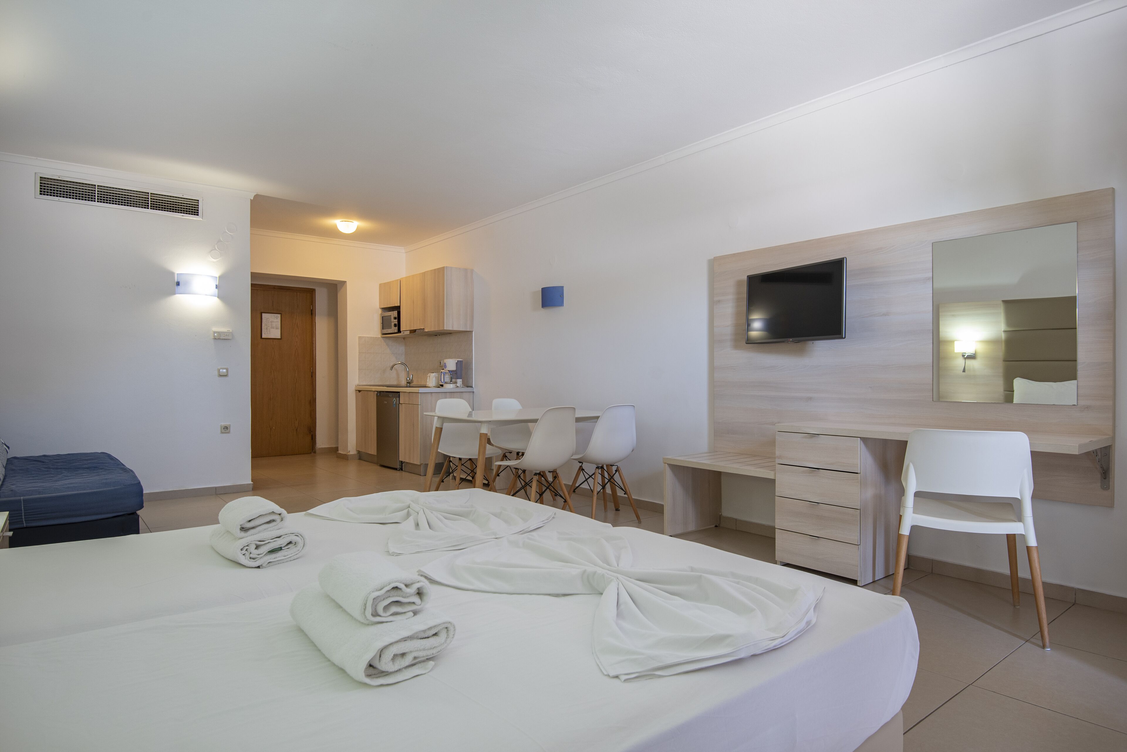 Quarto de hotel com cama, escrivaninha, televisão, espelho e cozinha compacta.