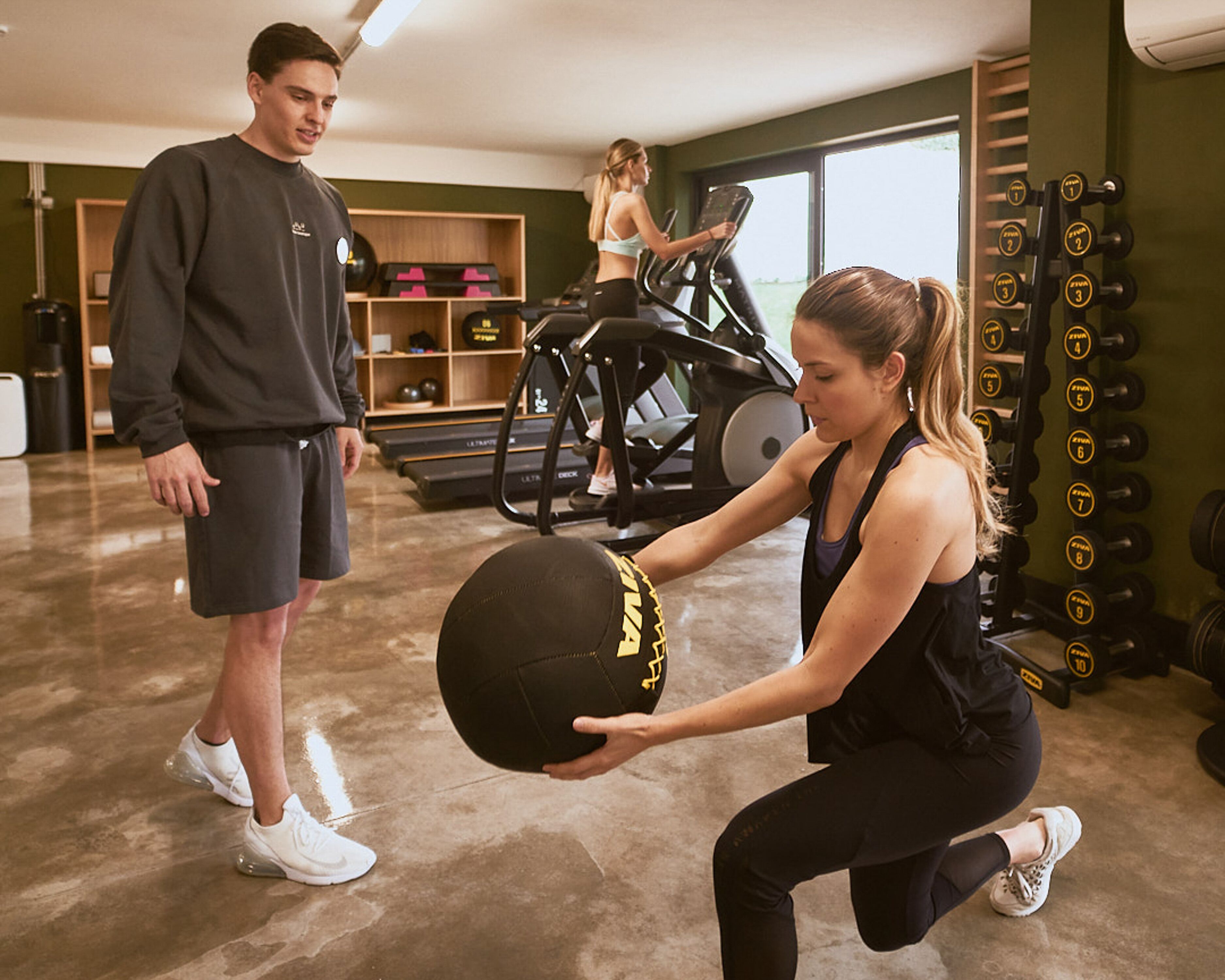 Uma academia com equipamentos de ginástica, esteira e uma pessoa segurando uma bola medicinal.