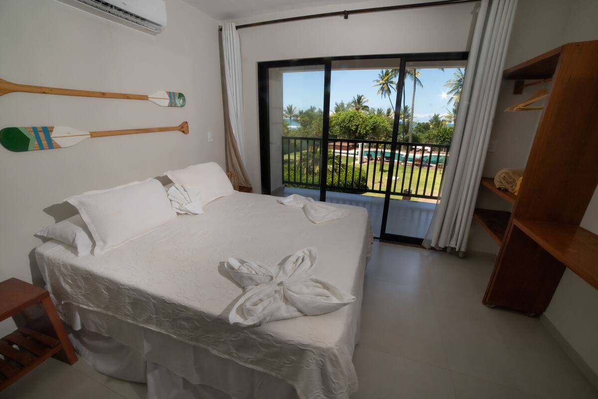 Quarto de hotel com uma cama, prateleira de madeira, janela com vista para uma piscina e palmeiras, e dois pranchas de remo na parede.