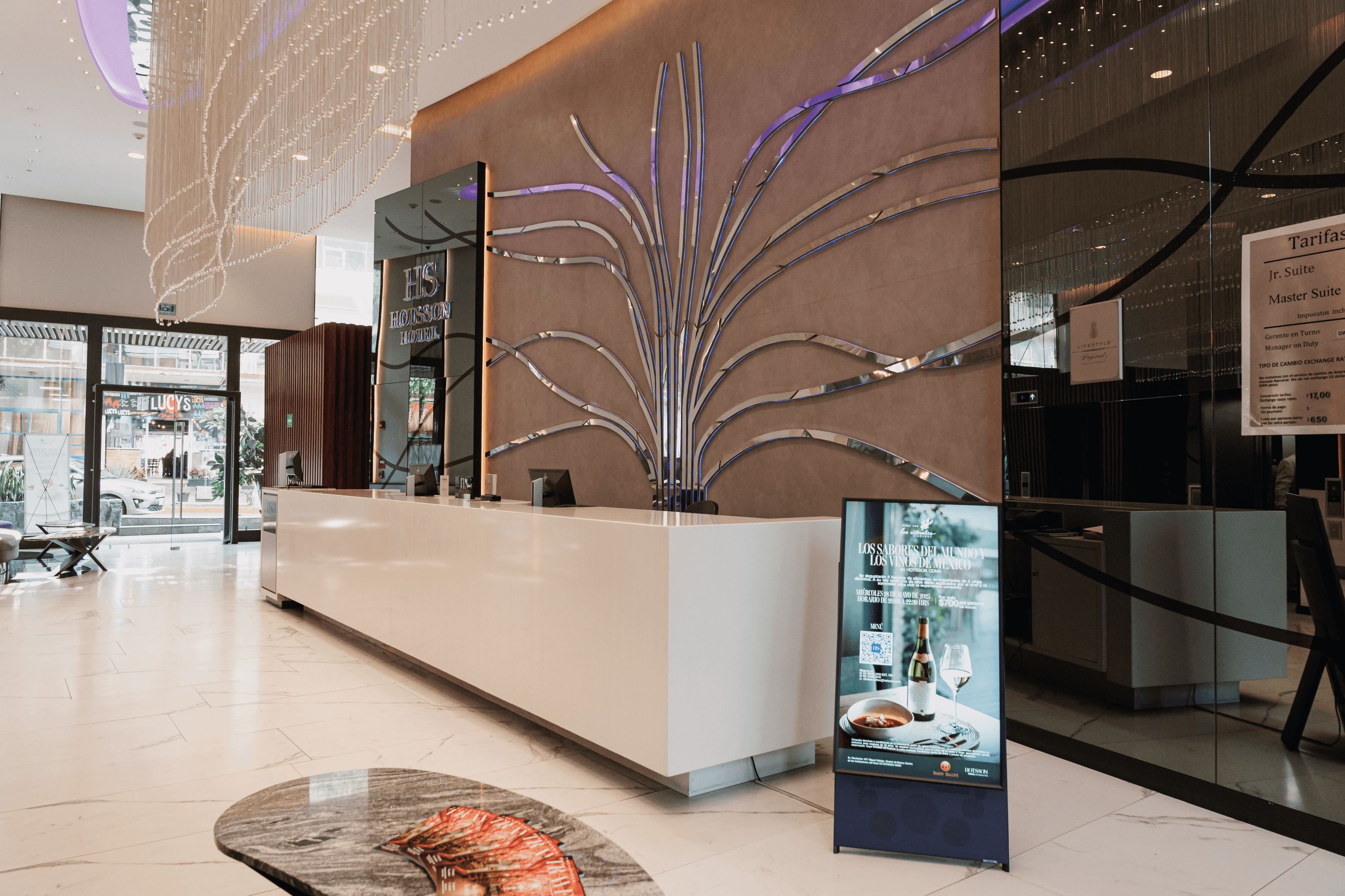Uma área de recepção de hotel moderna, com um balcão central, um detalhe decorativo na parede e um painel de informações digital.