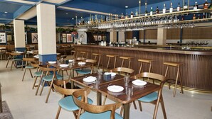 Restaurante com várias mesas e cadeiras, área de bar com diversos frascos e copos, e um interior limpo e moderno.