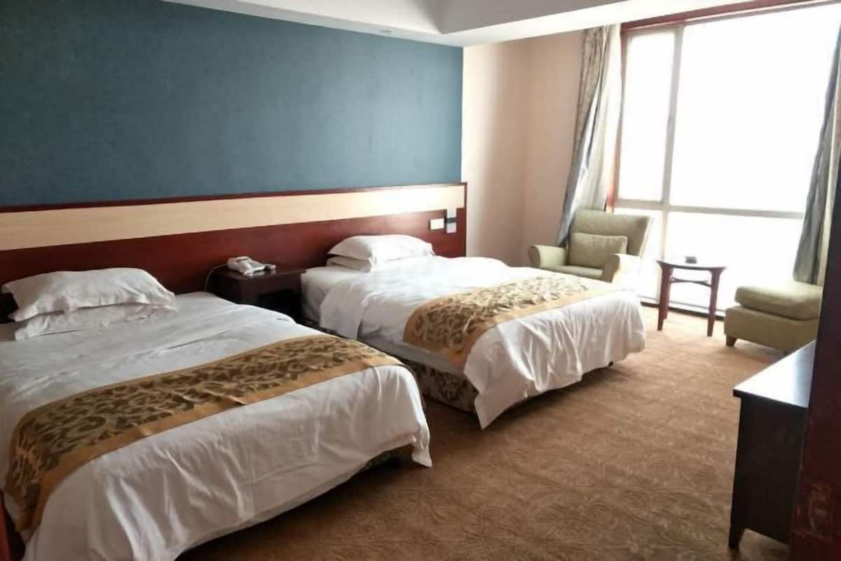 Quarto de hotel com duas camas, uma cadeira, uma mesinha e uma janela grande.