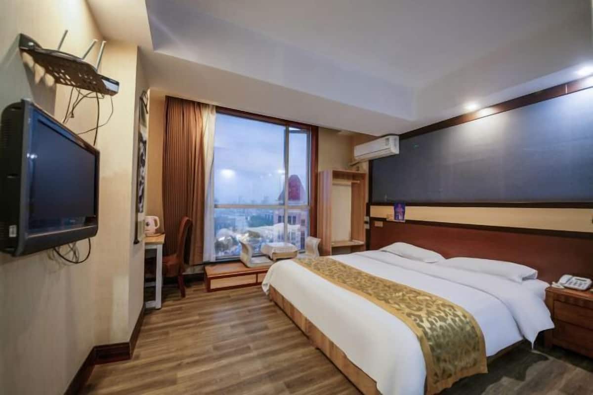 Quarto de hotel com uma cama grande, televisão, escrivaninha com cadeira, janela com cortinas e ar condicionado.
