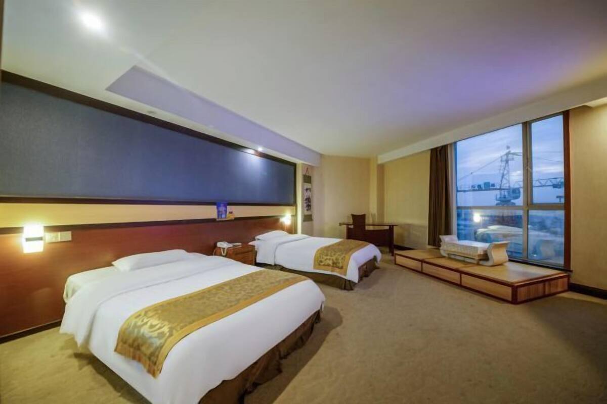 Quarto de hotel com duas camas, uma escrivaninha, uma cadeira e uma janela grande com vista para uma grua.