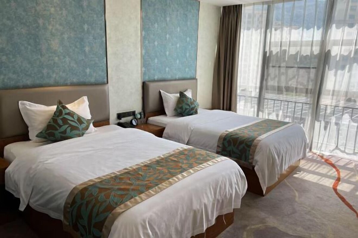 Duas camas lado a lado, com cabeceiras iguais, uma mesinha de cabeceira com relógio e vista para uma varanda por cortinas leves.
