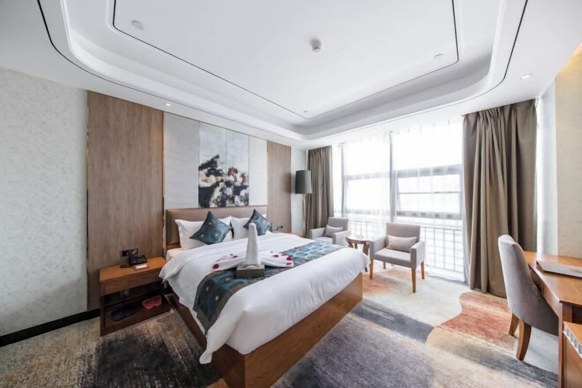 Quarto de hotel moderno com uma cama grande, uma escrivaninha e uma cadeira. Há um quadro na parede e uma janela com cortinas.